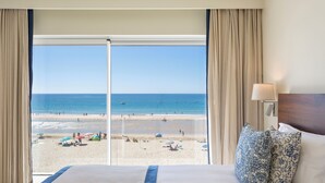 Quarto de hotel com uma janela ampla que oferece vista para a praia, uma cama com almofadas estampadas e uma luminária de cabeceira.