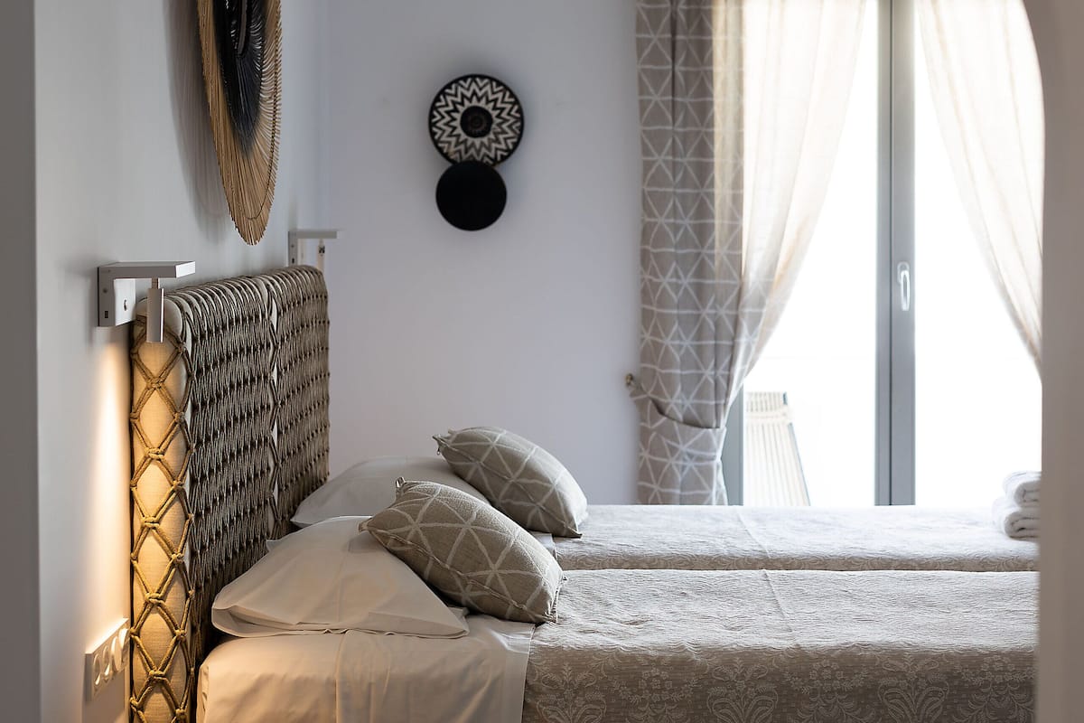 Quarto com cama, um banquinho de madeira, espelho fixado na parede e janela com cortinas.