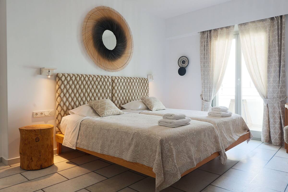 Quarto com cama, mesas de cabeceira, um espelho redondo e vista para o exterior através das cortinas.