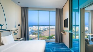 Quarto de hotel com uma cama grande, uma escrivaninha com cadeira, televisão e vista para a cidade e colinas.