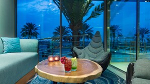 Quarto moderno com uma grande janela com vista para uma piscina e palmeiras. Há uma mesa de madeira redonda com bebidas e velas, um sofá e uma cadeira.
