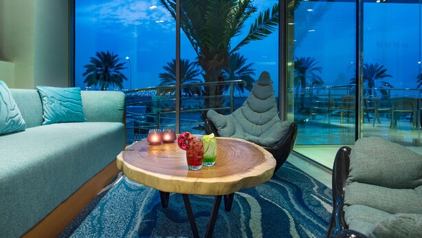 Uma área de estar moderna com sofá, mesa redonda de madeira com bebidas e uma janela ampla com vista para uma piscina e palmeiras.