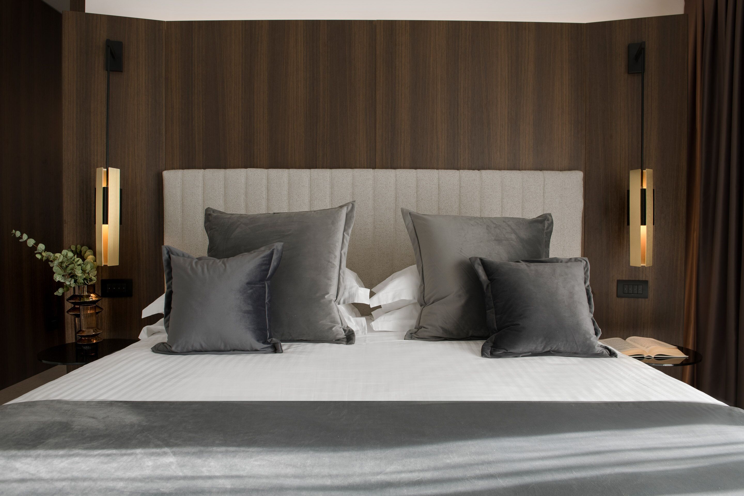 Quarto de hotel moderno com uma cama grande, duas luminárias de cabeceira e uma mesa de apoio com um livro e um vaso.