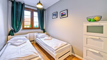 Quarto com duas camas de solteiro, um radiador, uma janela com cortinas, uma prateleira na parede com uma fruteira e duas imagens emolduradas.
