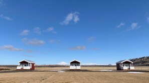 Duas pequenas cabanas térreas com exterior vermelho e branco, localizadas em uma paisagem plana e aberta, com montanhas distantes sob um céu azul límpido.