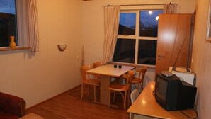 Um quarto pequeno com uma mesa e cadeiras de madeira, uma televisão e uma janela com cortinas.