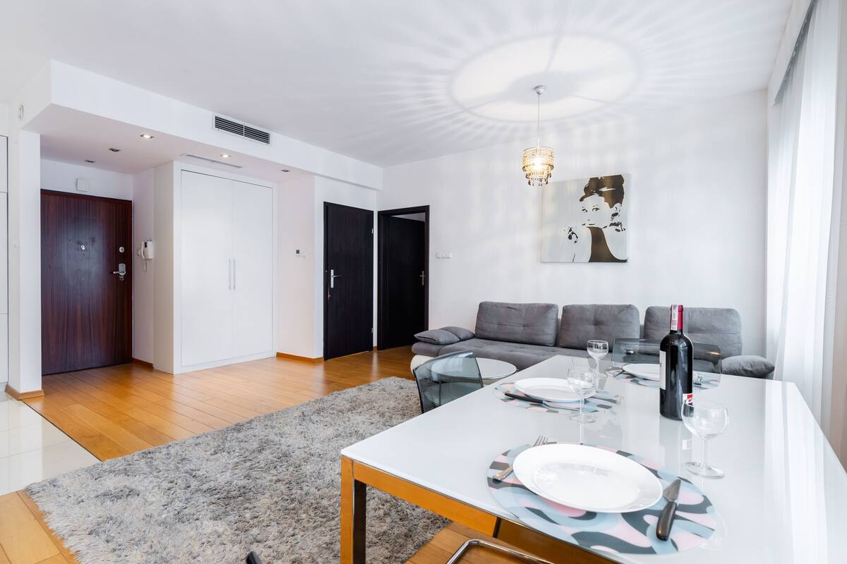 Uma sala de estar moderna com uma mesa de jantar posta para quatro pessoas, um sofá cinzento e um candelabro.