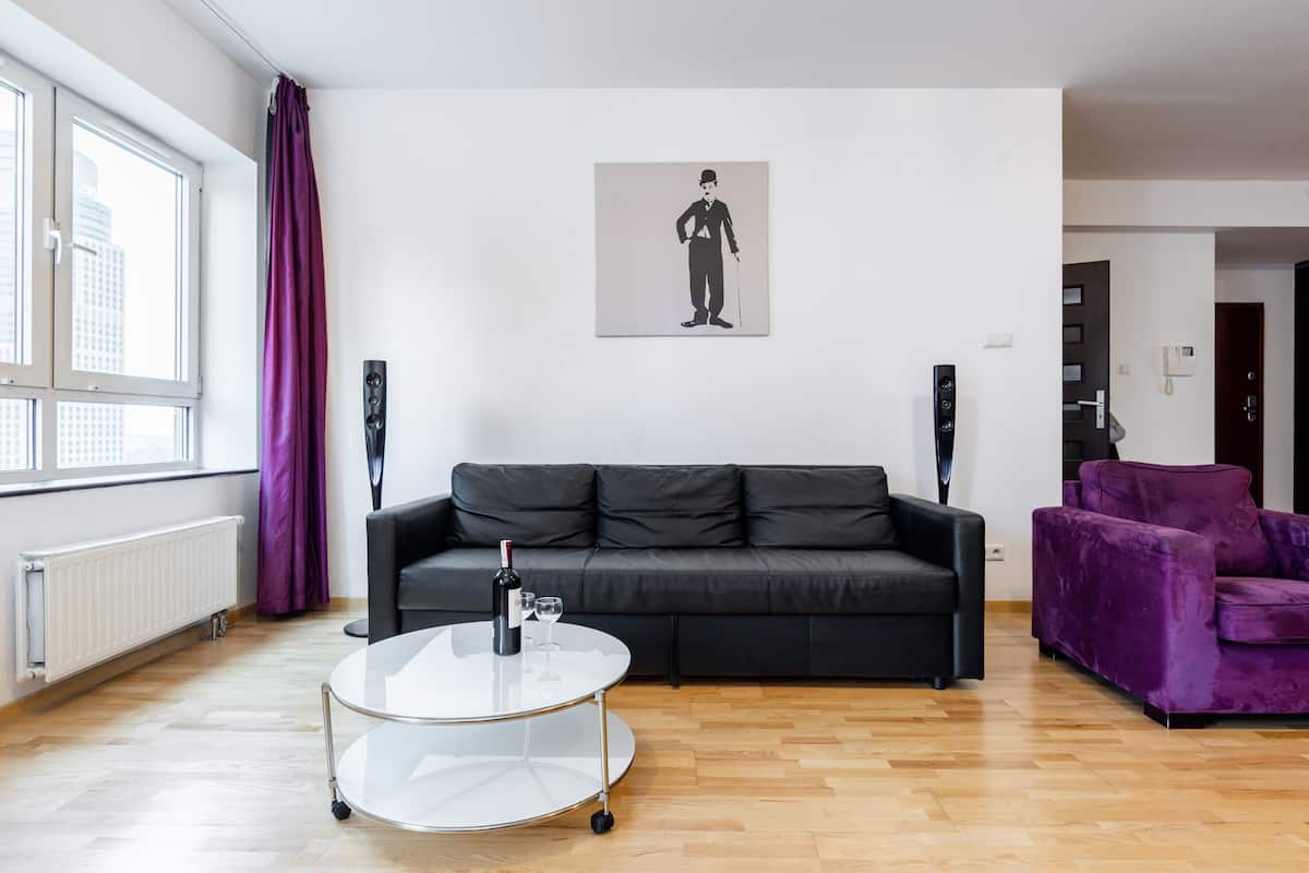 Uma sala de estar moderna com um sofá preto, uma mesa de centro branca e uma poltrona roxa. Há um quadro na parede e uma janela com cortinas.