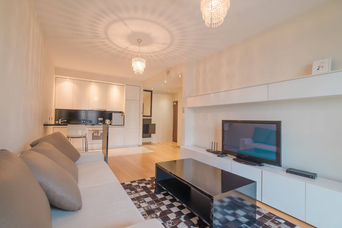 Uma sala de estar moderna com um sofá, uma mesa de centro e uma televisão. É visível uma cozinha ao fundo, com armários e uma área de jantar.