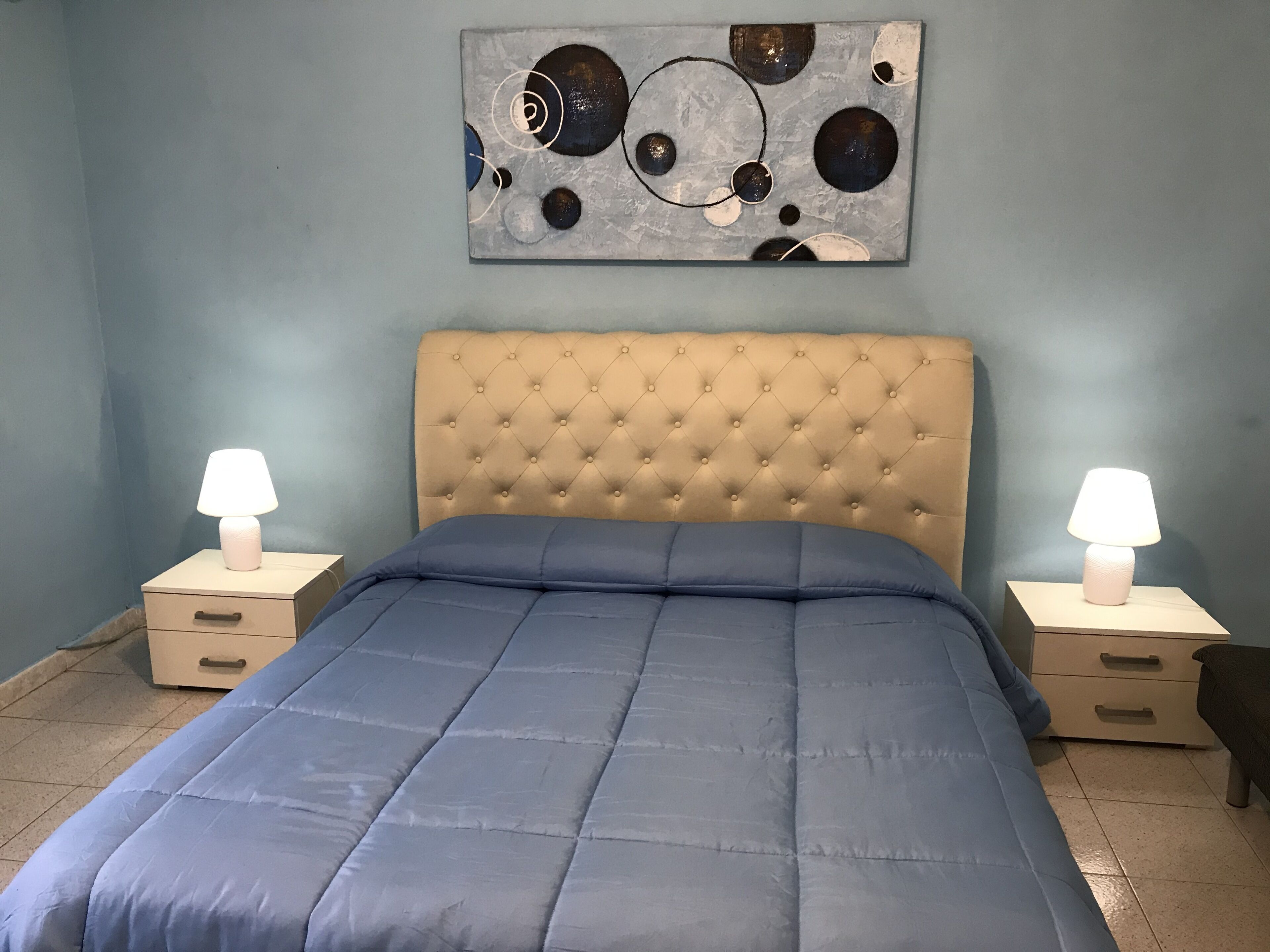 Quarto com cabeceira estofada, colcha azul, duas mesas de cabeceira com luminárias e uma obra de arte na parede acima da cama.