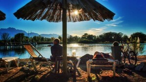 Um cenário à beira do lago com um guarda-sol de palha, cadeiras de descanso e uma bicicleta, transmitindo um espaço ao ar livre tranquilo para relaxamento.