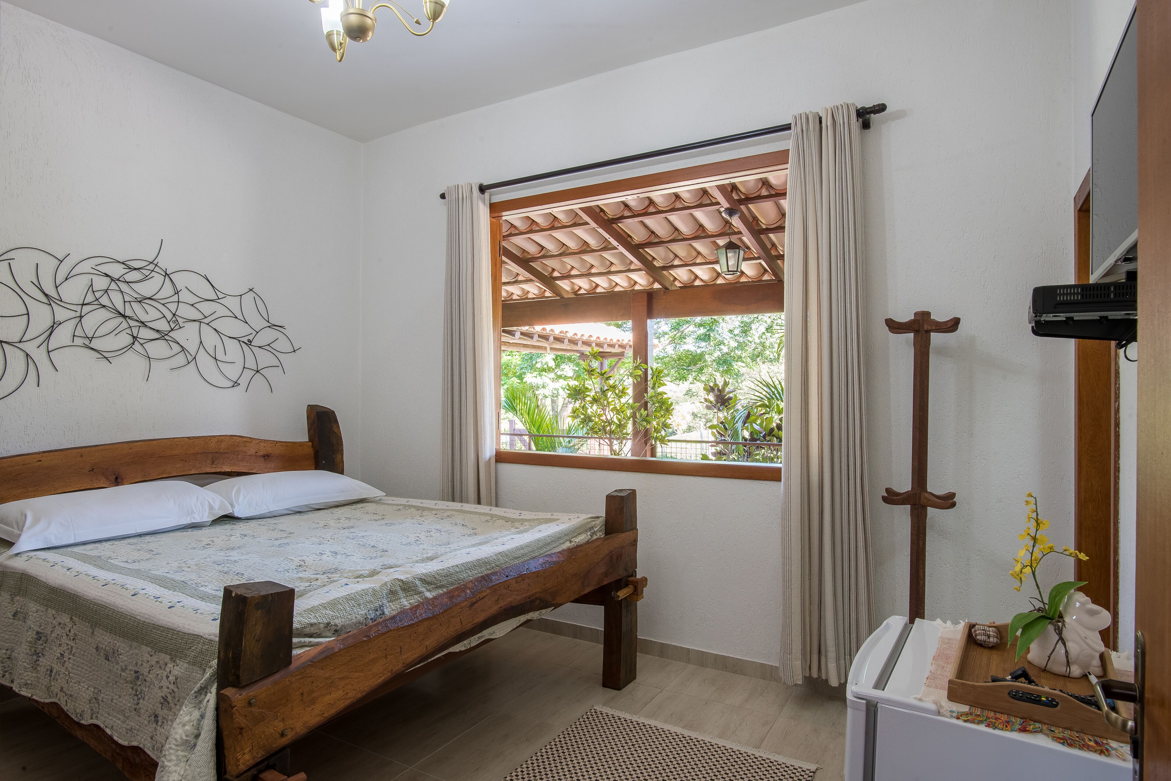 Quarto com cama de madeira, janela com vista para vegetação, uma obra de arte na parede e uma pequena mesa com uma planta.