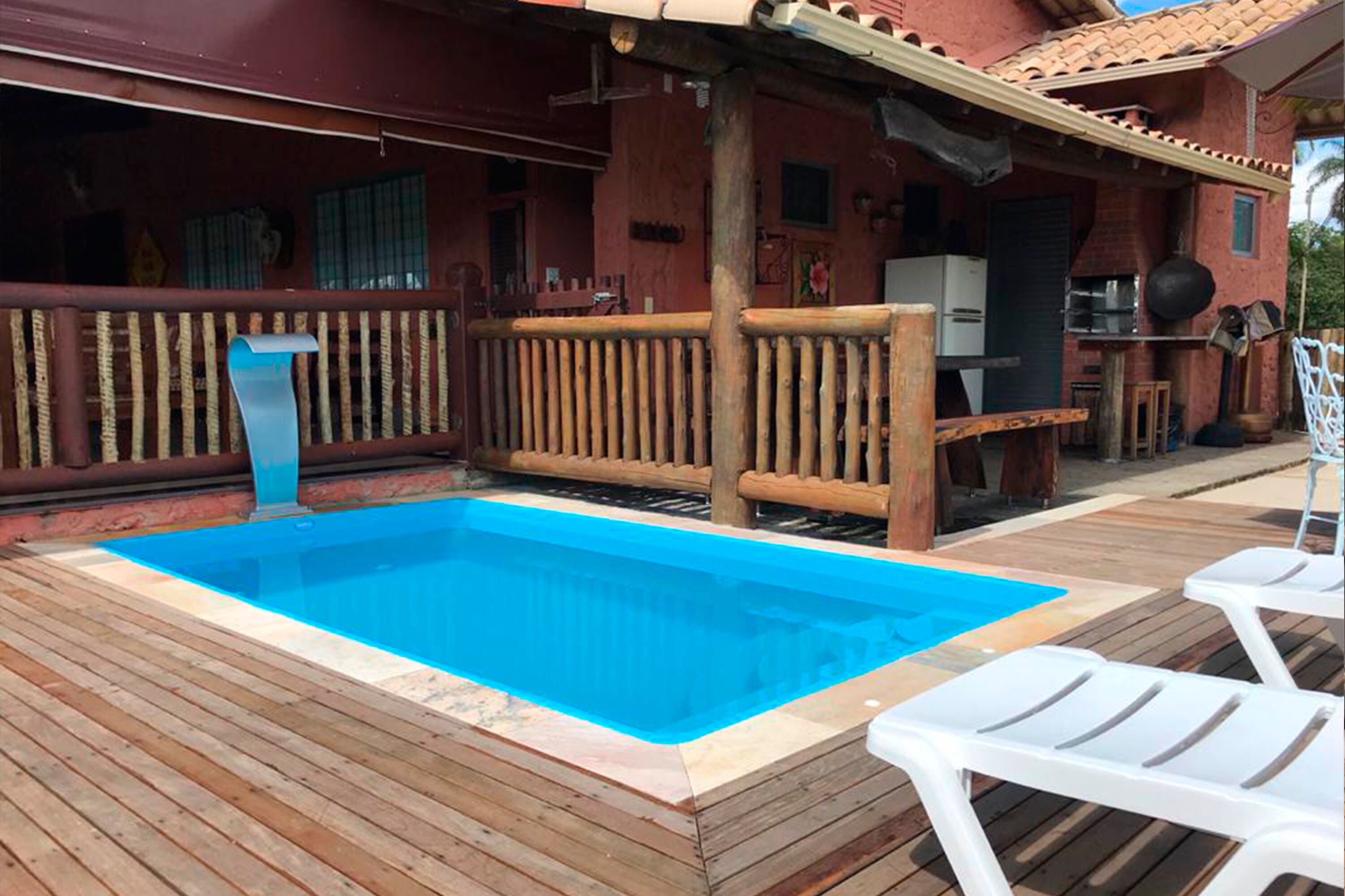 Uma piscina com deck de madeira, cerca de madeira e cadeiras de descanso brancas.