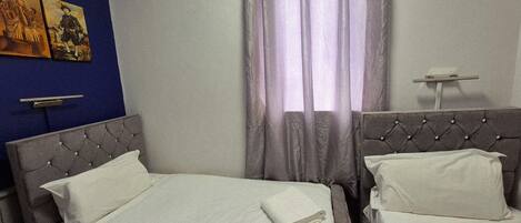 Quarto de hotel com duas camas, uma parede de destaque azul e uma janela com cortinas leves.