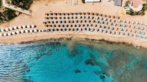 Vista aérea de uma praia com fileiras de espreguiçadeiras e guarda-sóis, mar azul cristalino e um pequeno edifício com telhado.