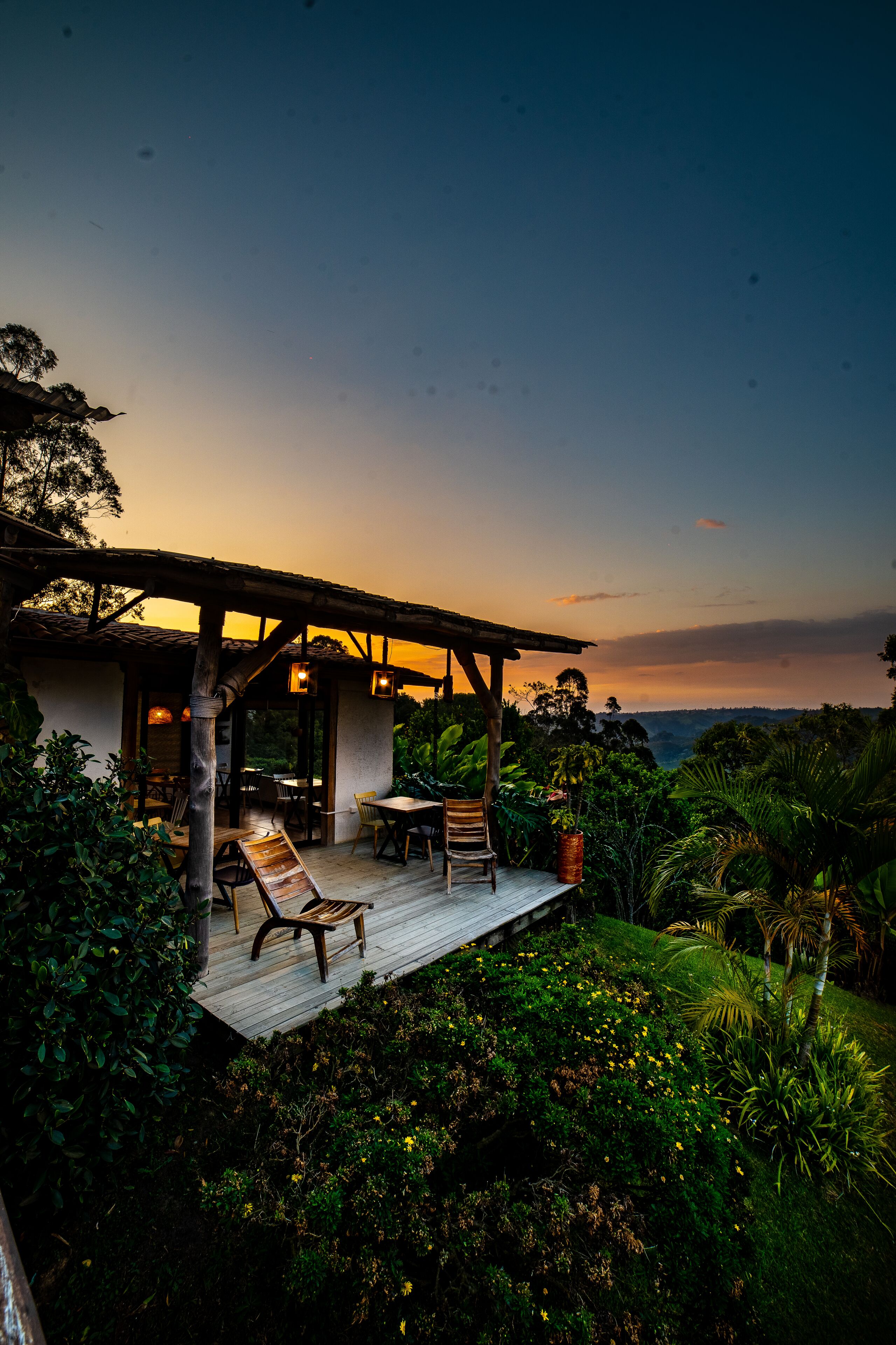 Um deck de madeira com mobiliário para área externa, cercado por vegetação exuberante e vista para a paisagem.