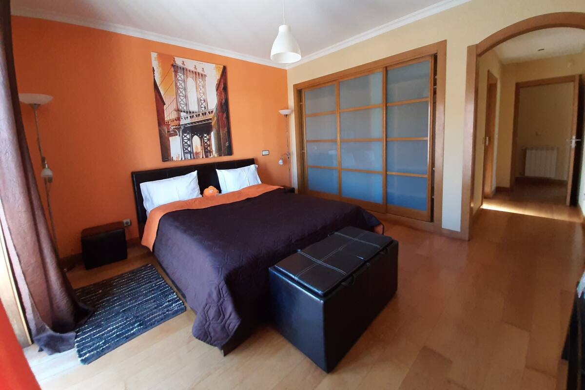Quarto com uma cama grande, mesa de cabeceira, um banquinho preto, um quadro na parede e uma porta de vidro deslizante que dá para outro cômodo.