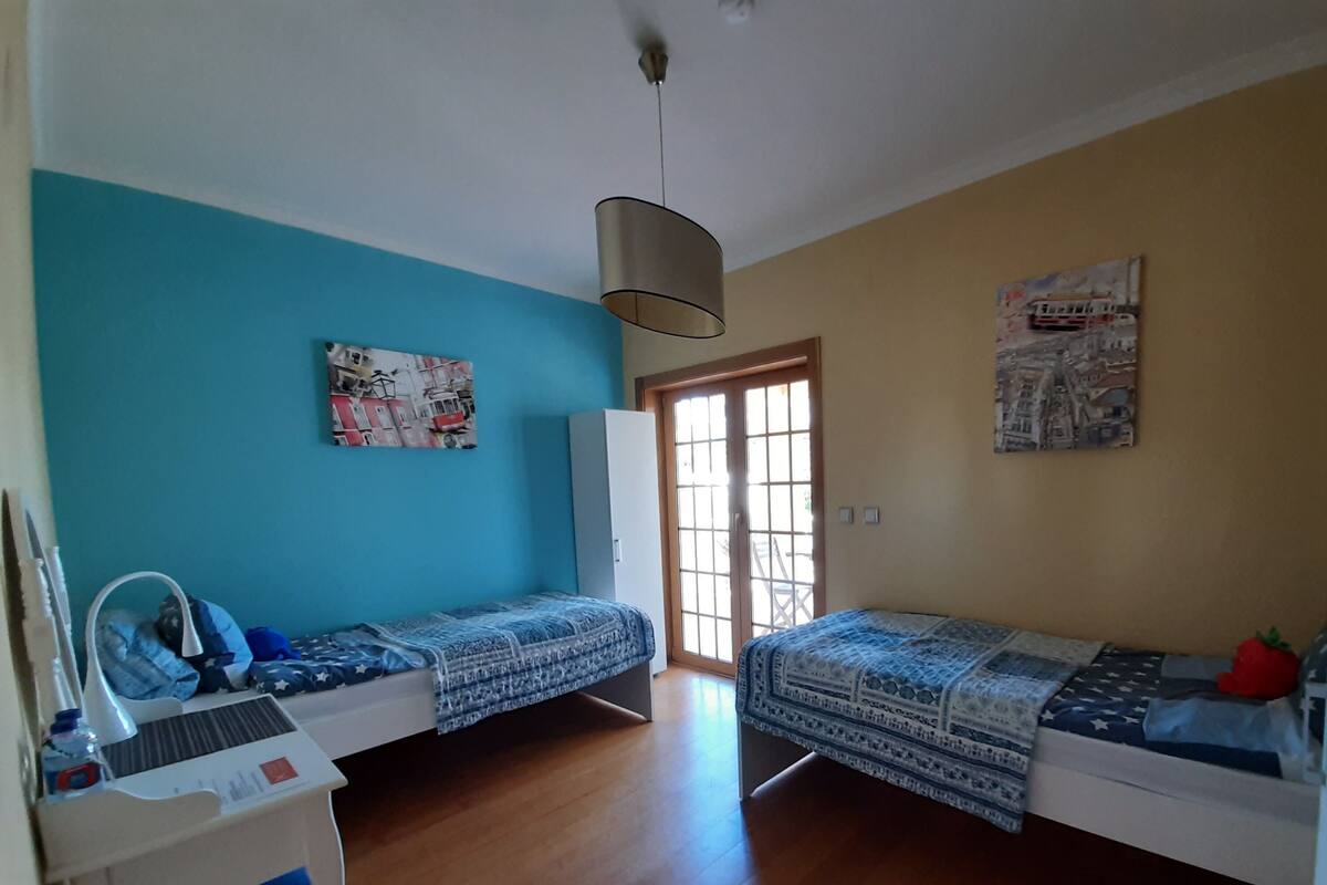 Quarto com duas camas de solteiro, uma escrivaninha, uma cadeira e uma janela.
