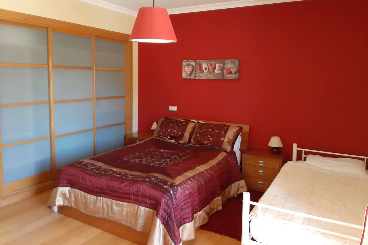 Quarto com cama, mesa de cabeceira, cômoda, parede vermelha e janela com persianas.