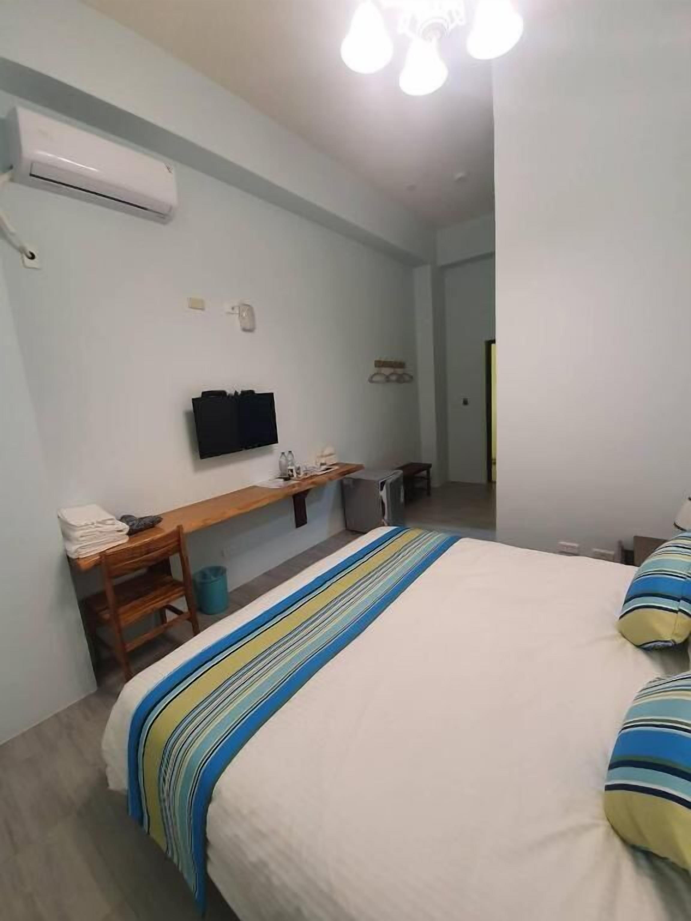 Quarto de hotel com cama, escrivaninha com televisão de tela plana, ar condicionado e uma pequena área de estar com cadeira e lixeira.