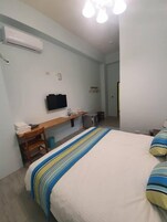 Quarto de hotel com cama, escrivaninha com televisão de tela plana, ar condicionado e uma pequena área de estar com cadeira e lixeira.