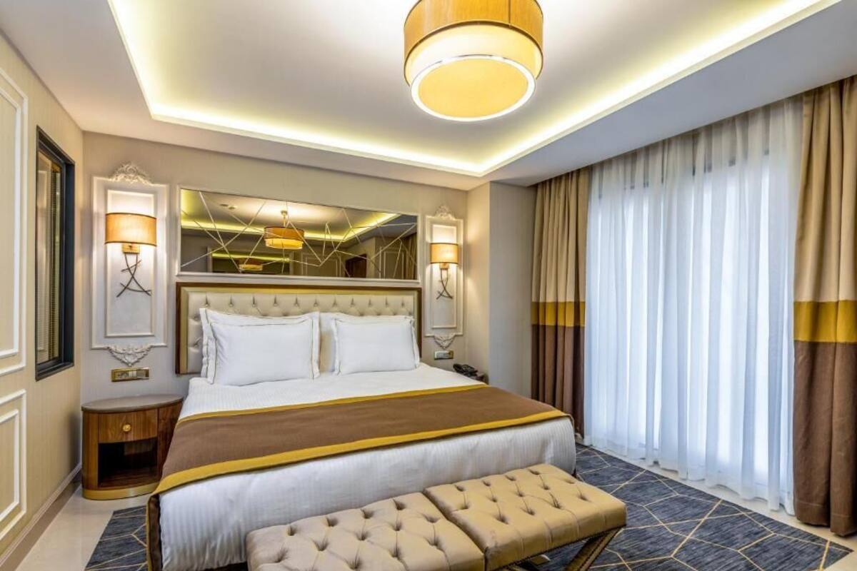 Quarto de hotel com uma cama grande, um banquinho estofado, um espelho, duas arandelas e uma janela ampla com cortinas.