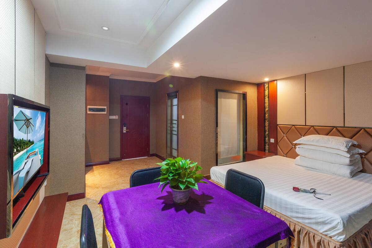 Quarto de hotel com cama, televisão, mesa com toalha de mesa roxa e um vaso de planta.