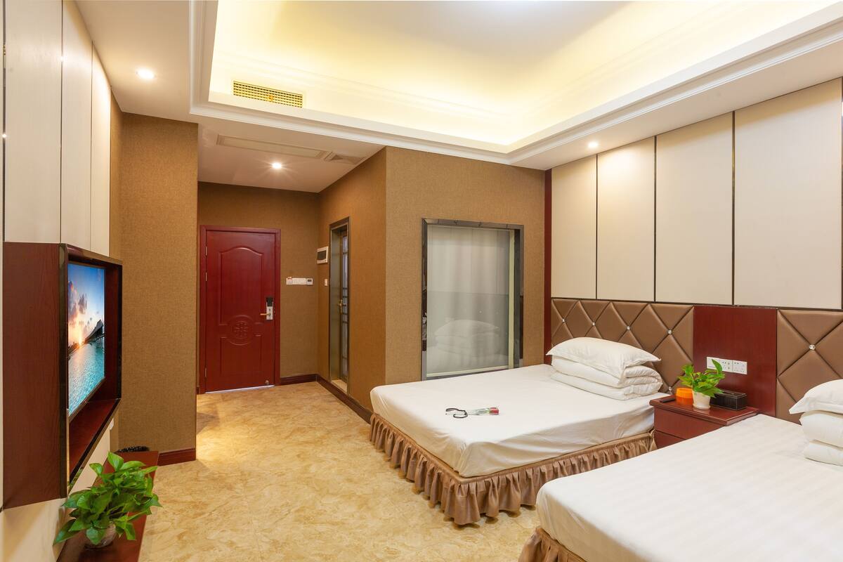 Quarto de hotel com duas camas, televisão, uma planta e porta para o banheiro.