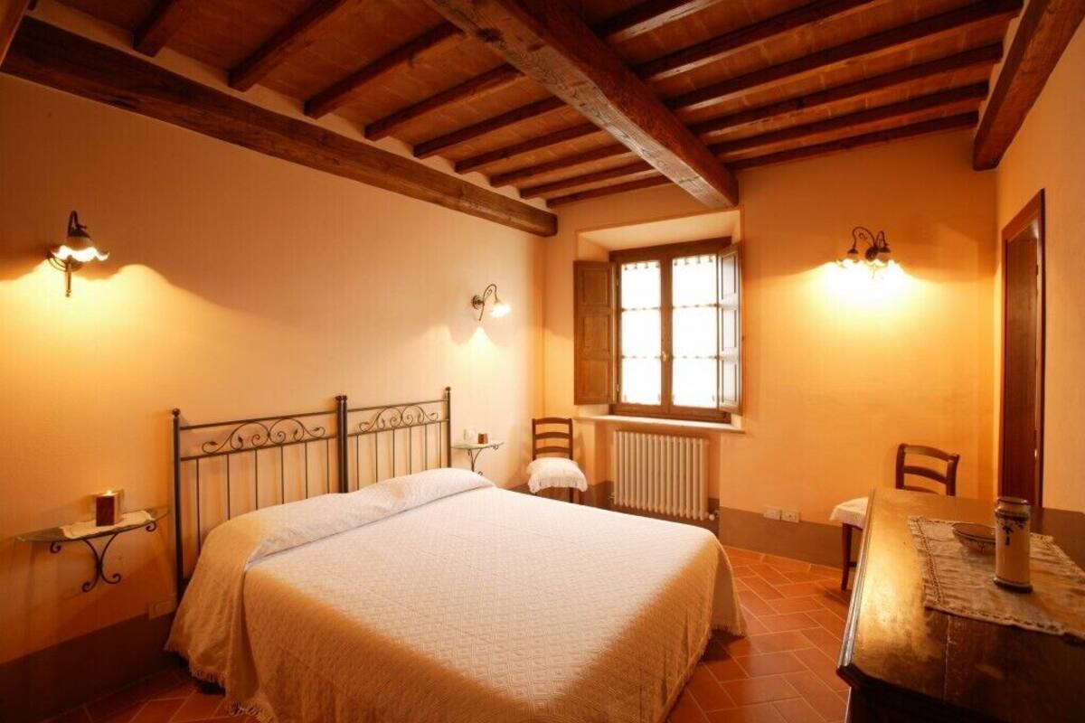 Quarto com cama, vigas de madeira no teto, janela com persianas e luminárias fixadas na parede.