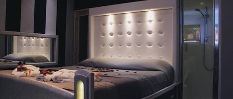 Quarto moderno com cabeceira estofada, box de vidro para o chuveiro e cama bem arrumada com toalhas.