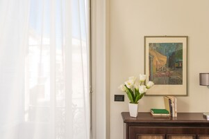 Um quarto com uma janela grande, um quadro na parede, um vaso branco com flores e um aparador de madeira com livros.