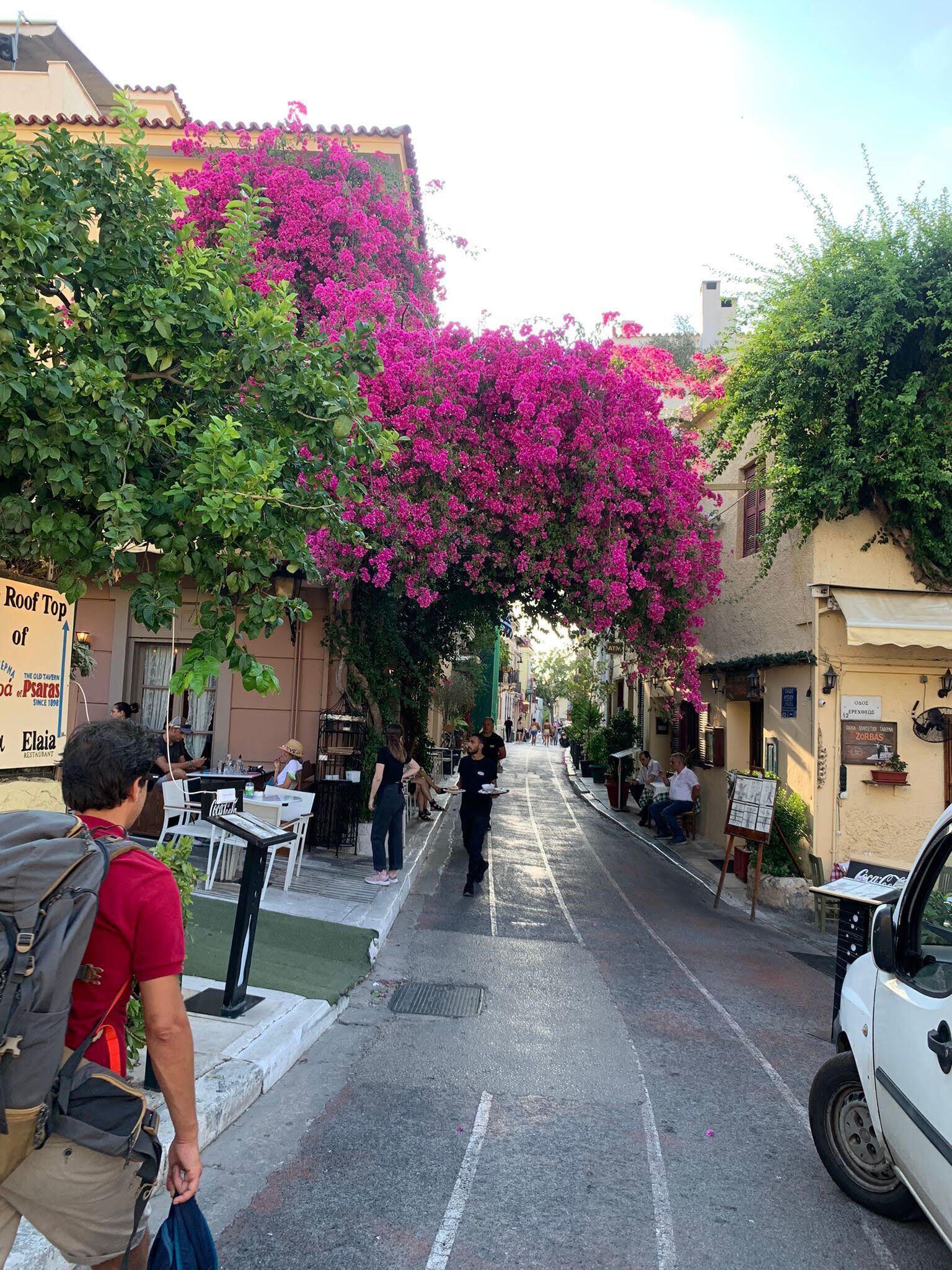 Rua com cafés e restaurantes, um homem caminhando com uma mochila e um carro estacionado.