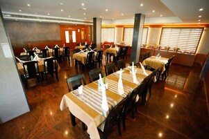 Uma ampla sala de jantar com várias mesas e cadeiras, piso lustroso e janelas grandes com persianas.
