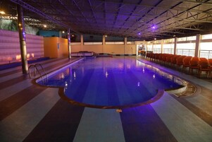 Piscina interna com superfície de água límpida, rodeada por piso e paredes revestidos, e teto com vigas aparentes.