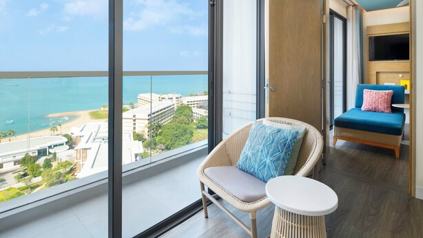 Uma varanda com vista para o mar e edifícios, cadeiras de vime, uma mesa pequena e um sofá azul com uma almofada rosa.