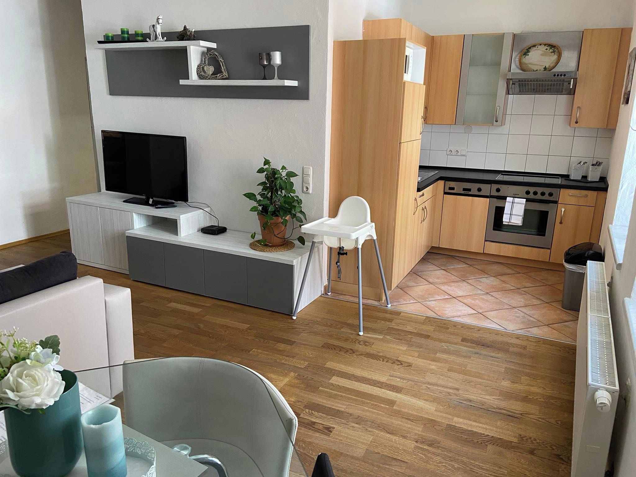 Cozinha moderna com fogão, forno e lava-louças. Possui área de jantar com mesa e cadeiras, e sala de estar com televisão e estantes.