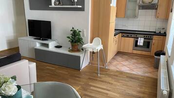 Cozinha moderna com fogão, forno e lava-louças. Possui área de jantar com mesa e cadeiras, e sala de estar com televisão e estantes.