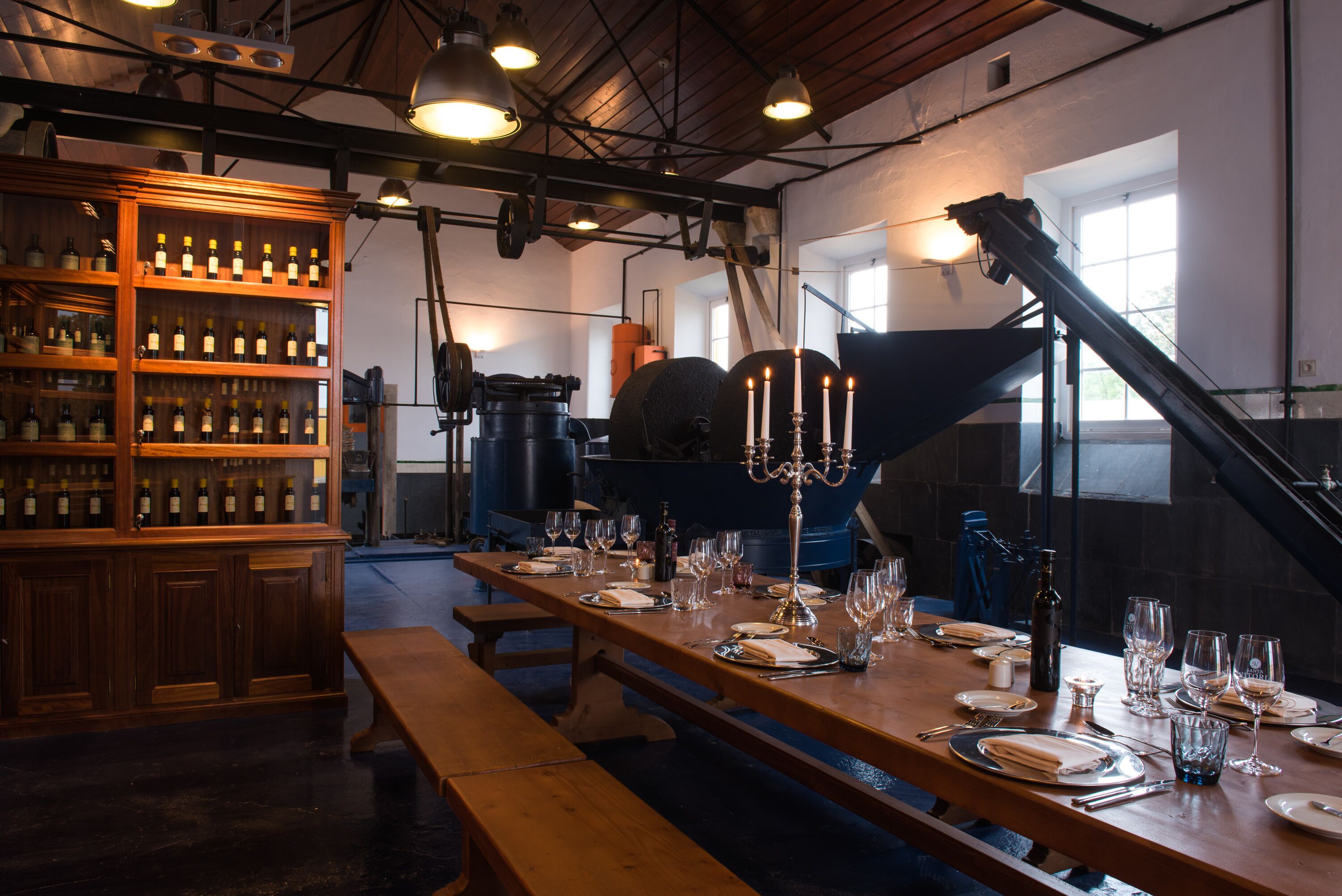 Uma sala de jantar com uma longa mesa de madeira posta para refeição, um aparador de vinhos à esquerda e uma escada ao fundo.