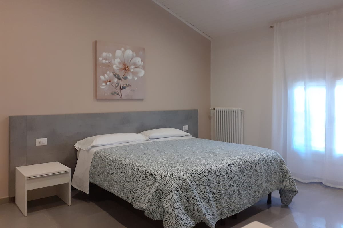 Quarto com cama, mesa de cabeceira, radiador e janela com cortinas.
