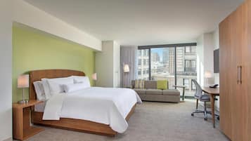 Quarto de hotel moderno com uma cama grande, um sofá, uma mesa de escritório e vista da cidade por grandes janelas.