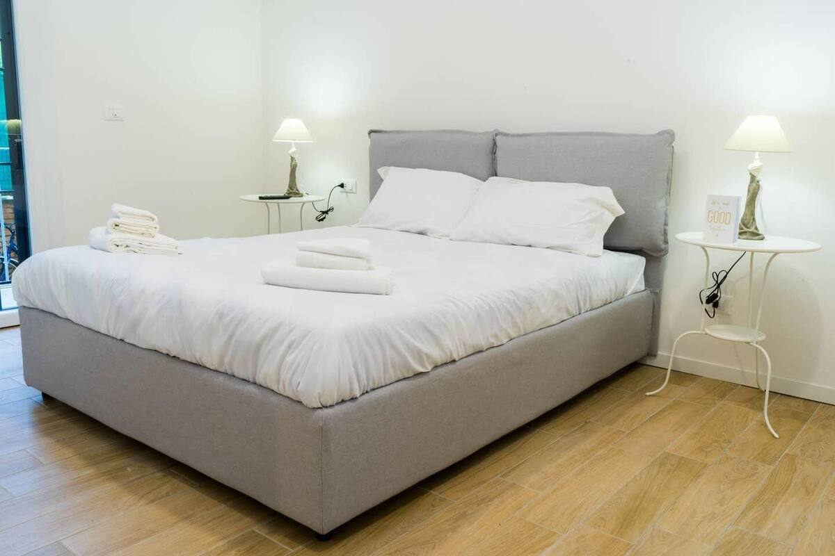 Uma cama bem arrumada com lençóis brancos e toalhas dobradas, uma cabeceira estofada cinza e duas mesas de cabeceira com luminárias e uma placa com a palavra 'GOOD'.