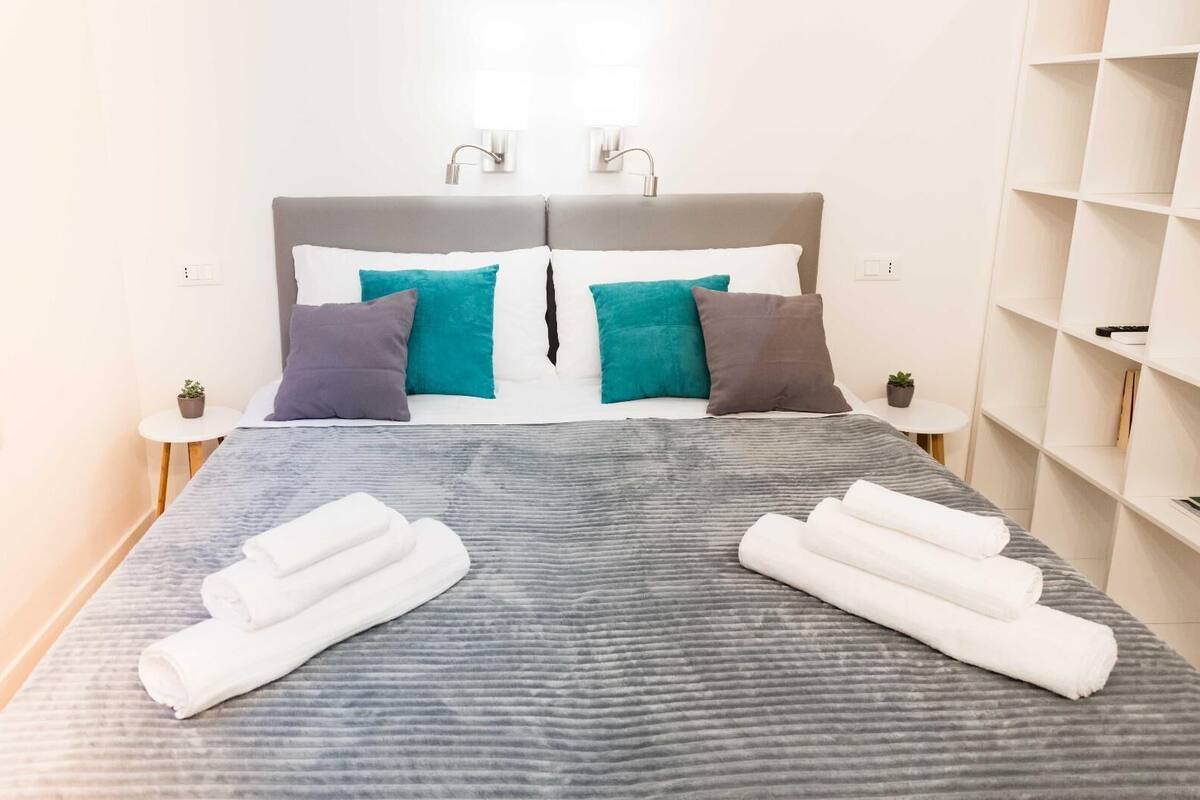 Cama bem arrumada com colcha cinza listrada, dois travesseiros brancos e dois travesseiros verde-água. Duas mesinhas de cabeceira com pequenas plantas em vasos.