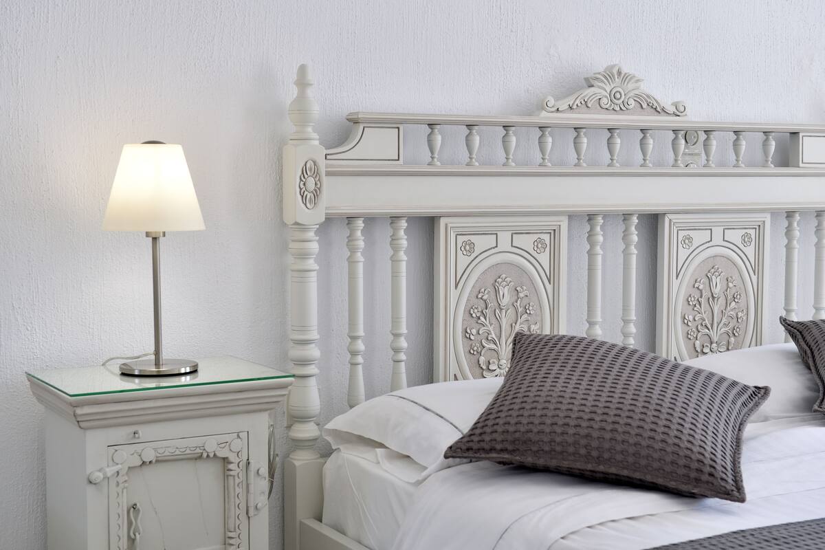 Uma cama bem arrumada com lençóis brancos e almofadas decorativas, uma mesinha de cabeceira branca com luminária, e uma cabeceira branca com esculturas detalhadas.