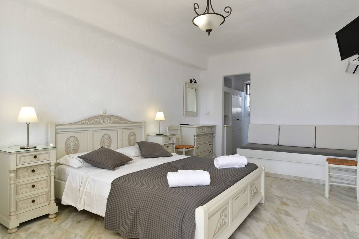 Quarto com cama, mesas de cabeceira, um banquinho, um espelho e uma TV fixada na parede.