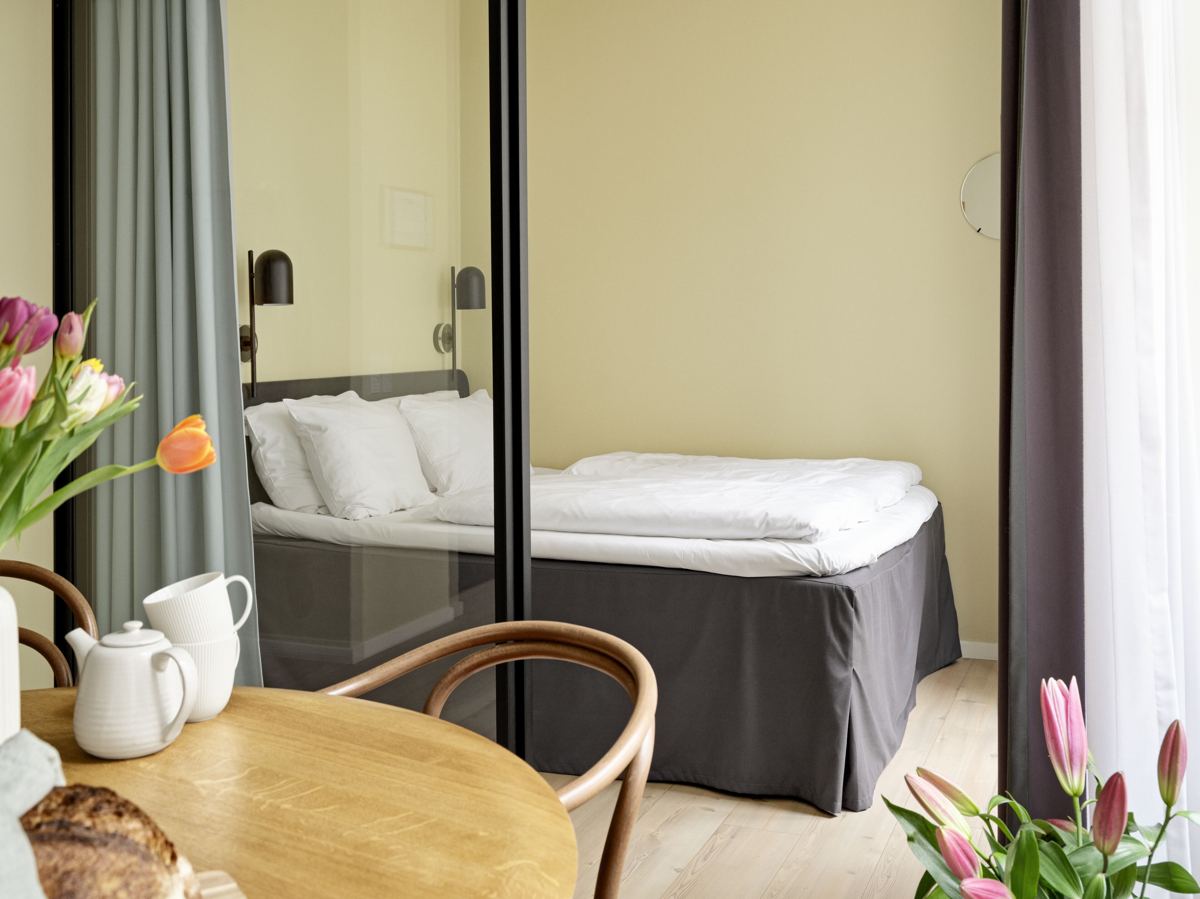 Quarto de hotel com uma cama, uma mesa de madeira com um jarro branco e uma xícara, e uma cadeira.