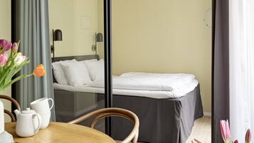 Quarto de hotel com uma cama, uma mesa de madeira com um jarro branco e uma xícara, e uma cadeira.