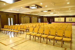 Uma sala de conferências com fileiras de cadeiras amarelas, um palco com uma mesa vermelha e ventiladores de teto.