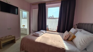 Quarto com cama, mesa de cabeceira, televisão na parede e janela com cortinas.