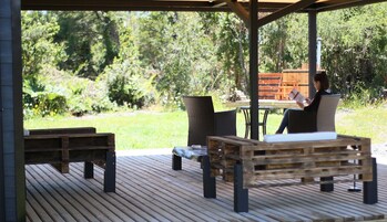Um deck de madeira ao ar livre com área de estar coberta, incluindo uma mesa e cadeiras, com vista para uma área gramada com árvores.