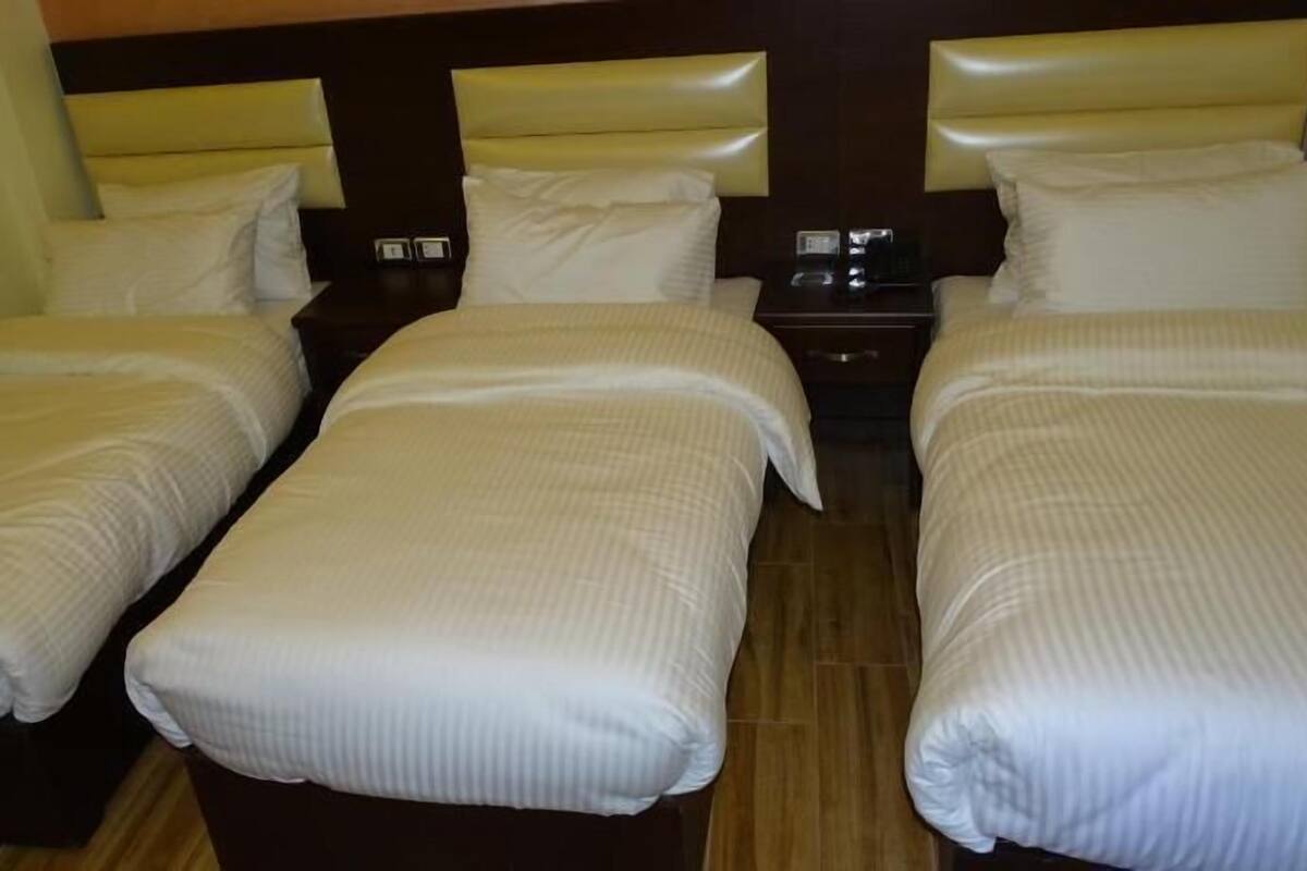 Quarto de hotel com três camas de solteiro, cada uma com cabeceira e travesseiro. Um criado-mudo entre as camas possui um telefone e uma pequena mesa.